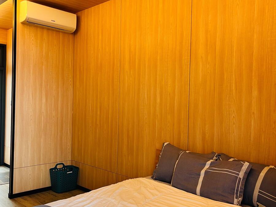 Quarto de hotel com parede de madeira, cama com colcha branca e cinza, duas almofadas, um cesto de cor escura e um aparelho de ar condicionado.