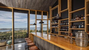 Uma área de bar com prateleiras de madeira repletas de garrafas, um vaso de vidro grande e vista para um lago.
