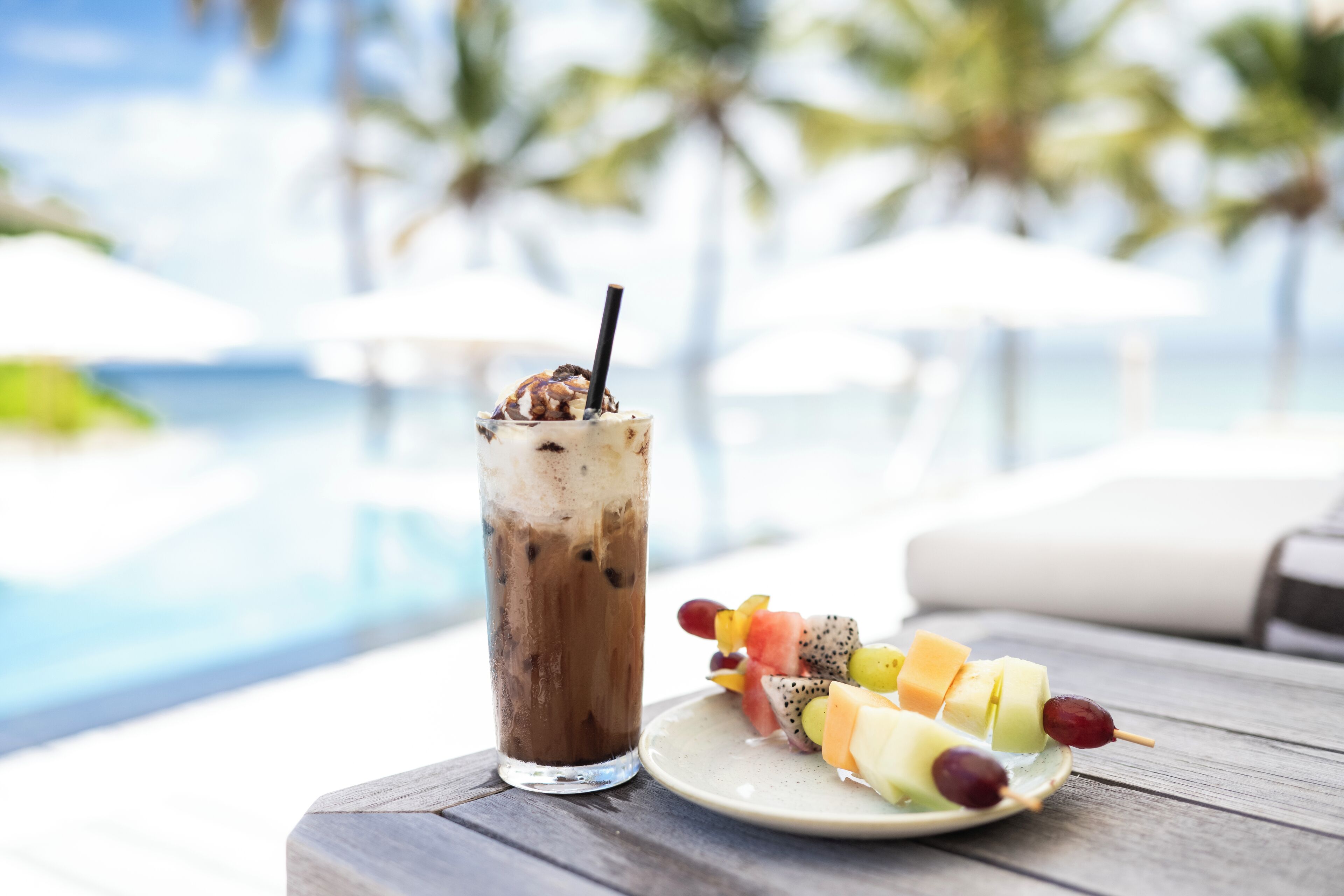 Uma bebida e um espeto de frutas sobre uma mesa de madeira perto de uma piscina, com vista para palmeiras e um corpo d'água.