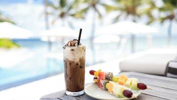 Uma bebida e um espeto de frutas sobre uma mesa de madeira perto de uma piscina, com vista para palmeiras e um corpo d'água.