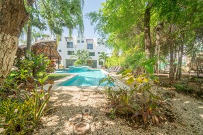 Uma área de piscina com estrutura de telhado de palha, rodeada por vegetação exuberante e um edifício branco ao fundo.