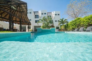Piscina com água azul cristalina, cercada por um edifício moderno com varandas e uma estrutura de telhado de palha.