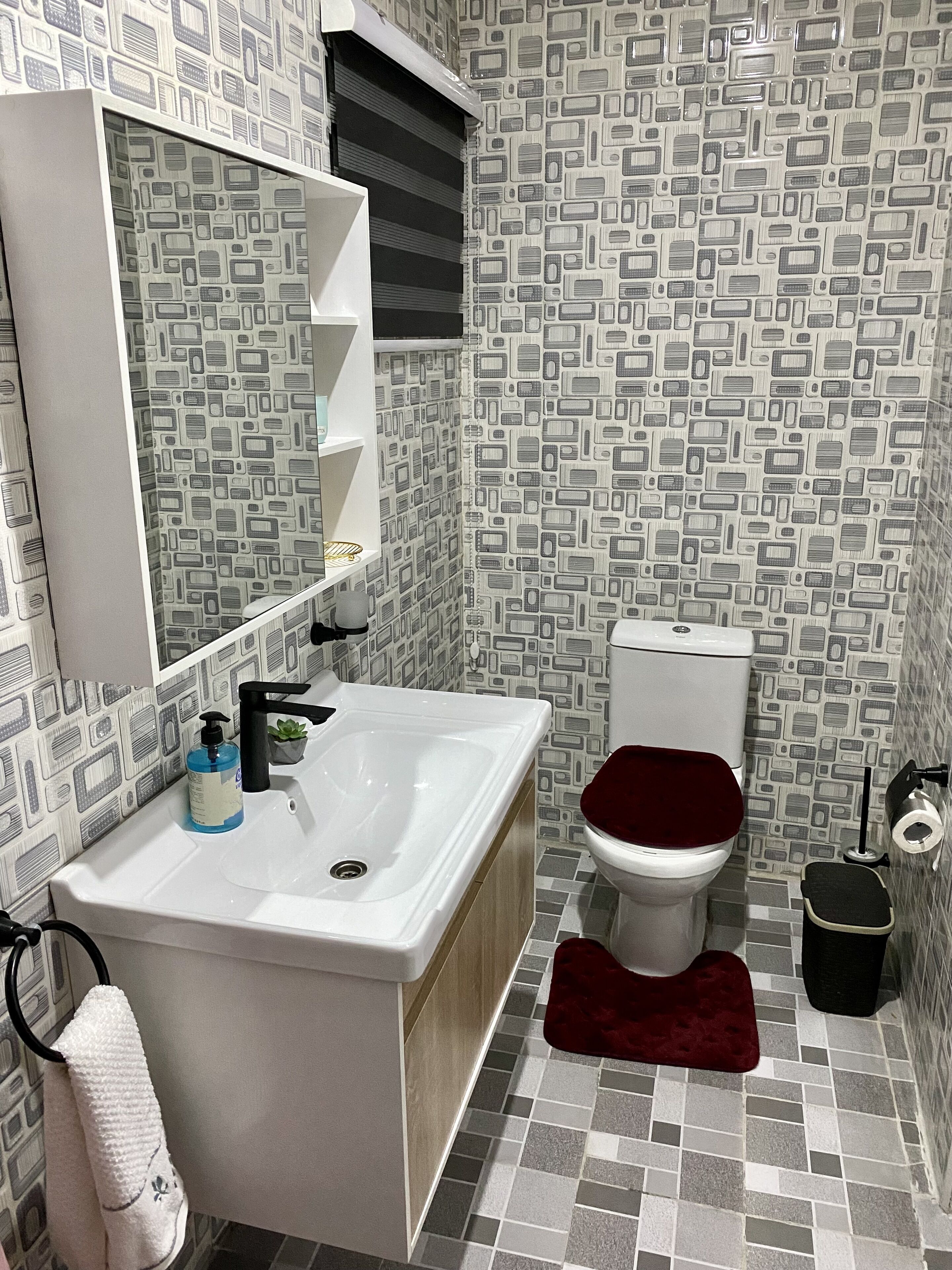 Uma casa de banho com uma pia branca, um espelho, uma sanita com assento vermelho e azulejos estampados nas paredes e no chão.