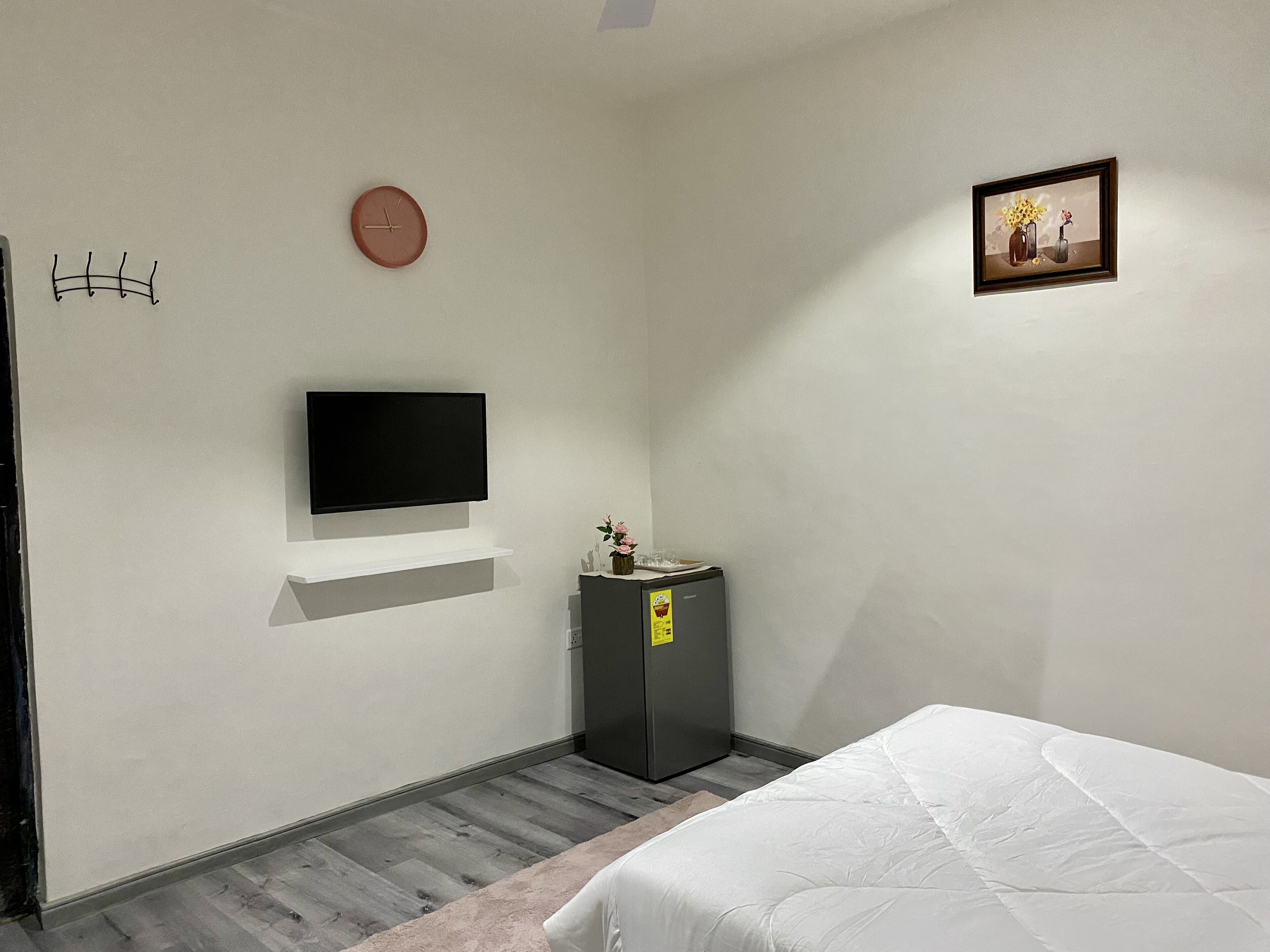 Quarto com cama, televisão na parede, pequena geladeira, relógio e quadro emoldurado.