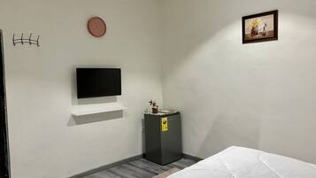 Quarto com cama, televisão na parede, pequena geladeira, relógio e quadro emoldurado.