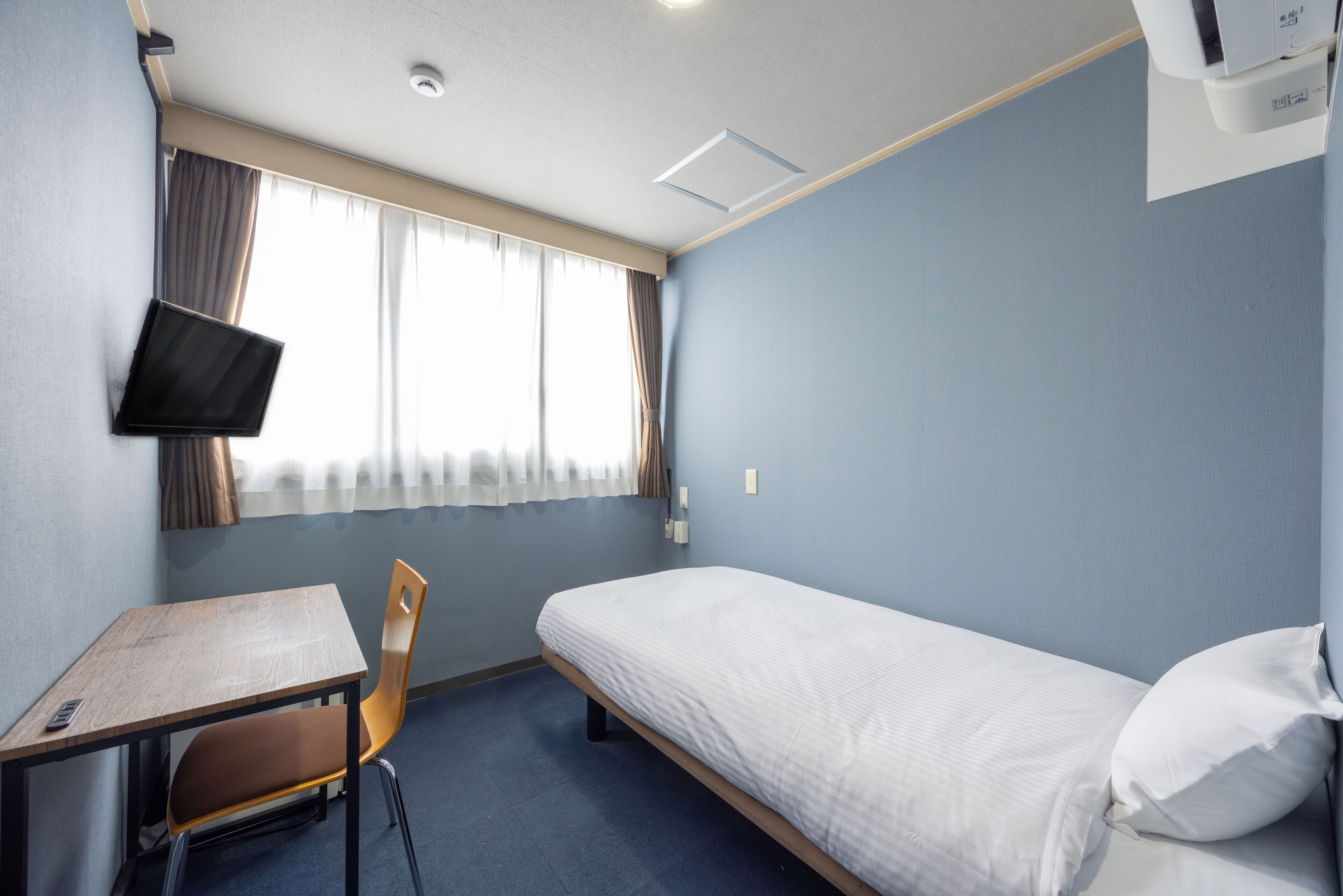 Quarto de hotel com uma cama de solteiro, uma escrivaninha, uma cadeira, televisão de tela plana na parede e uma janela com cortinas.