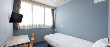 Quarto de hotel com uma cama de solteiro, uma escrivaninha, uma cadeira, televisão de tela plana na parede e uma janela com cortinas.