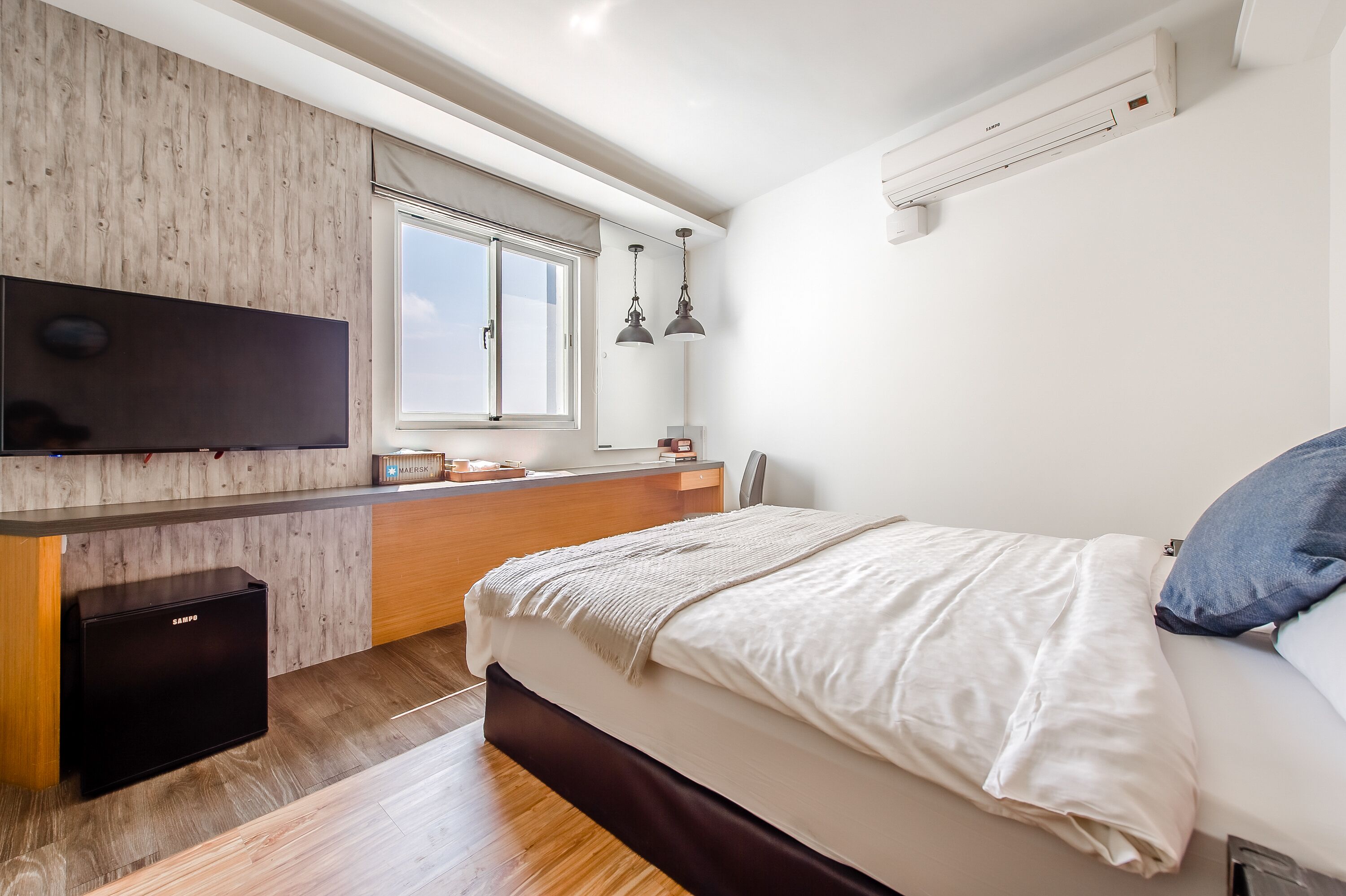 Um quarto moderno com piso de madeira, uma cama com roupa de cama branca, uma televisão montada na parede, uma janela com vista e um pequeno armário preto.