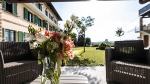 Uma mesa de vime com um vaso de flores, duas cadeiras de vime e vista para um edifício e uma área gramada.