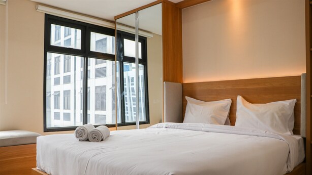 Cama bem arrumada com lençóis e almofadas brancos, cabeceira de madeira e uma janela grande com vista para edifícios.