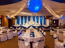 Uma sala de banquetes com mesas redondas dispostas para um evento formal, cobertas com toalhas azuis e brancas, e uma grande decoração floral azul pendurada no teto.