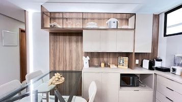 Cozinha compacta com micro-ondas, torradeira e cafeteira. Há uma mesa de jantar com cadeiras e um objeto decorativo sobre a mesa.