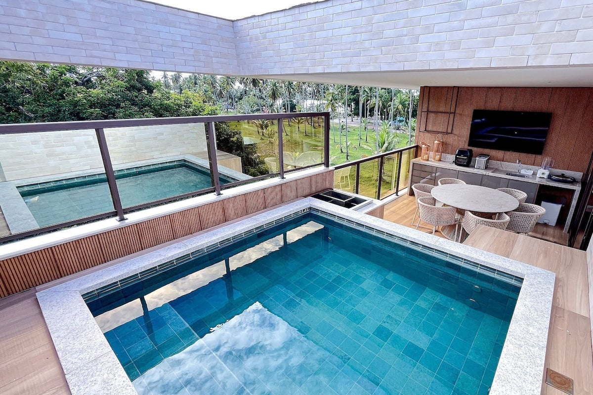 Uma piscina no terraço com guarda-corpo de vidro, área de jantar com móveis de vime e cozinha com televisão de tela plana.