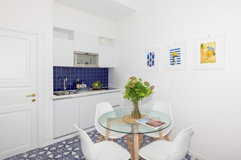 Uma cozinha moderna com armários brancos, painel de fundo azul e uma mesa de jantar de vidro com cadeiras brancas. Há um vaso com flores sobre a mesa.