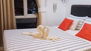 Quarto de hotel com cama, roupa de cama listrada, cabeceira preta e almofadas vermelhas e brancas.