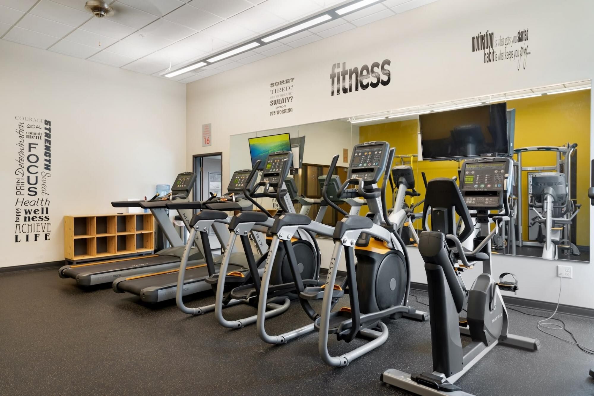 Centro de fitness com esteiras, elípticos e uma parede com frases motivacionais.