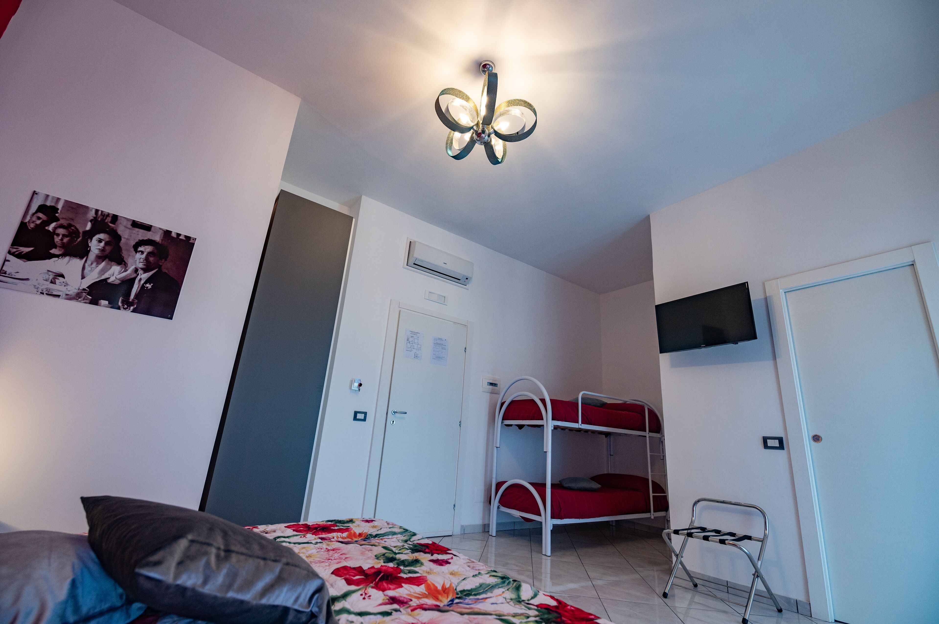 Quarto com beliches, cama com colcha florida, televisão fixada na parede e ar condicionado.