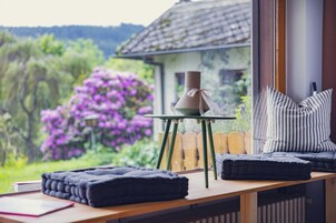 Quarto aconchegante com uma mesa de madeira, uma cadeira, uma mesinha com uma vela e vista para uma casa e jardim.