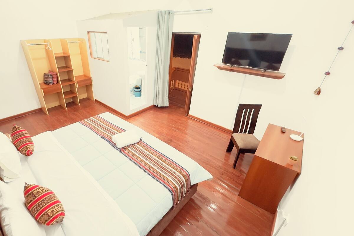 Quarto com cama, escrivaninha, televisão, guarda-roupa e banheiro visível por uma porta aberta.