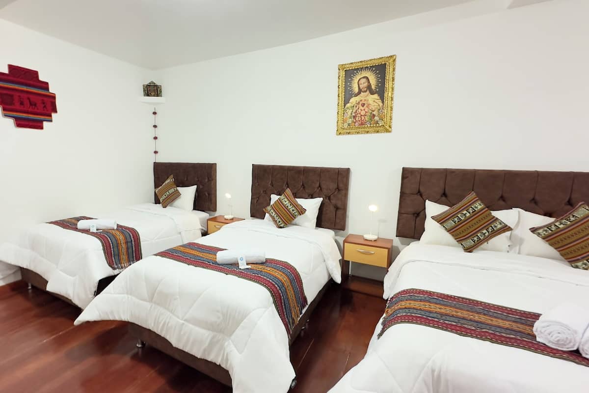 Quarto de hotel com três camas, cada uma com roupa de cama branca e almofadas decorativas. Há uma obra de arte na parede acima das camas e uma pequena mesa com abajur entre elas.