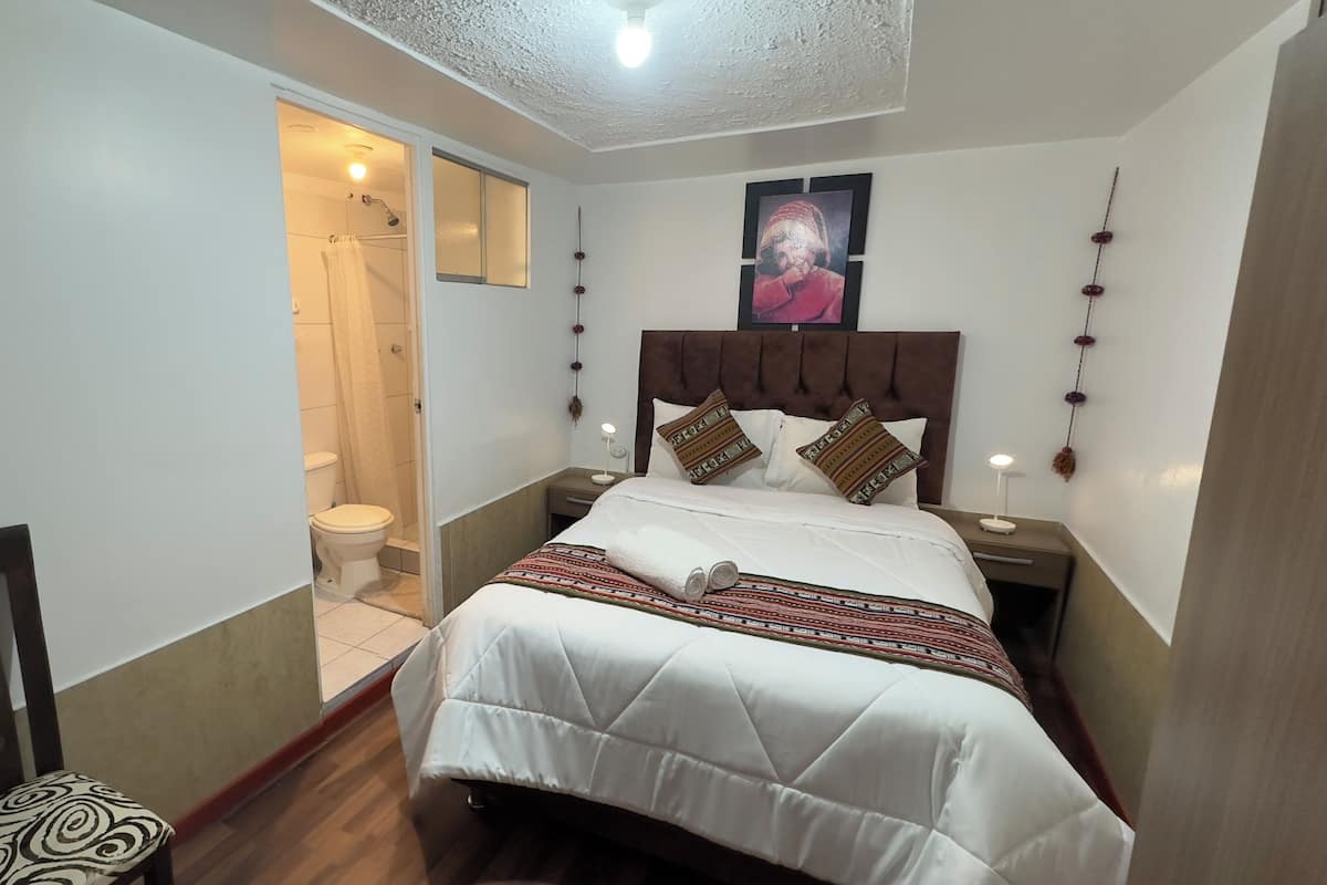 Quarto com cama, mesa de cabeceira, cadeira, banheiro com vaso sanitário e chuveiro, e uma luminária no teto.