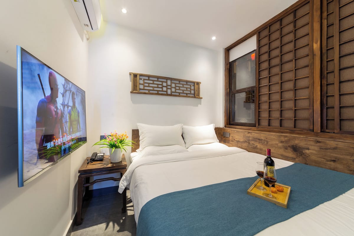 Quarto de hotel com cama, mesa de cabeceira com telefone e um vaso de flores, televisão na parede e janela com persiana de madeira.