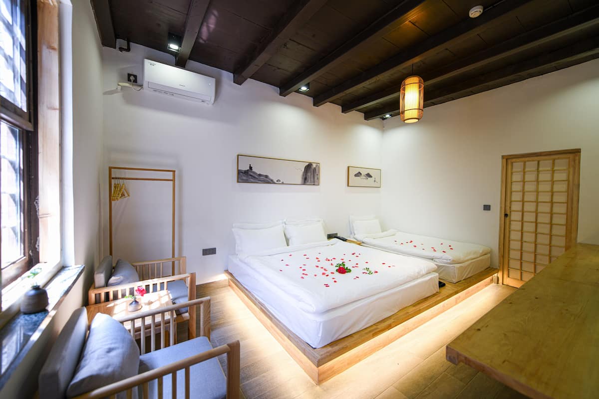 Quarto de hotel com duas camas, um banco de madeira, uma mesinha e uma janela com vista.