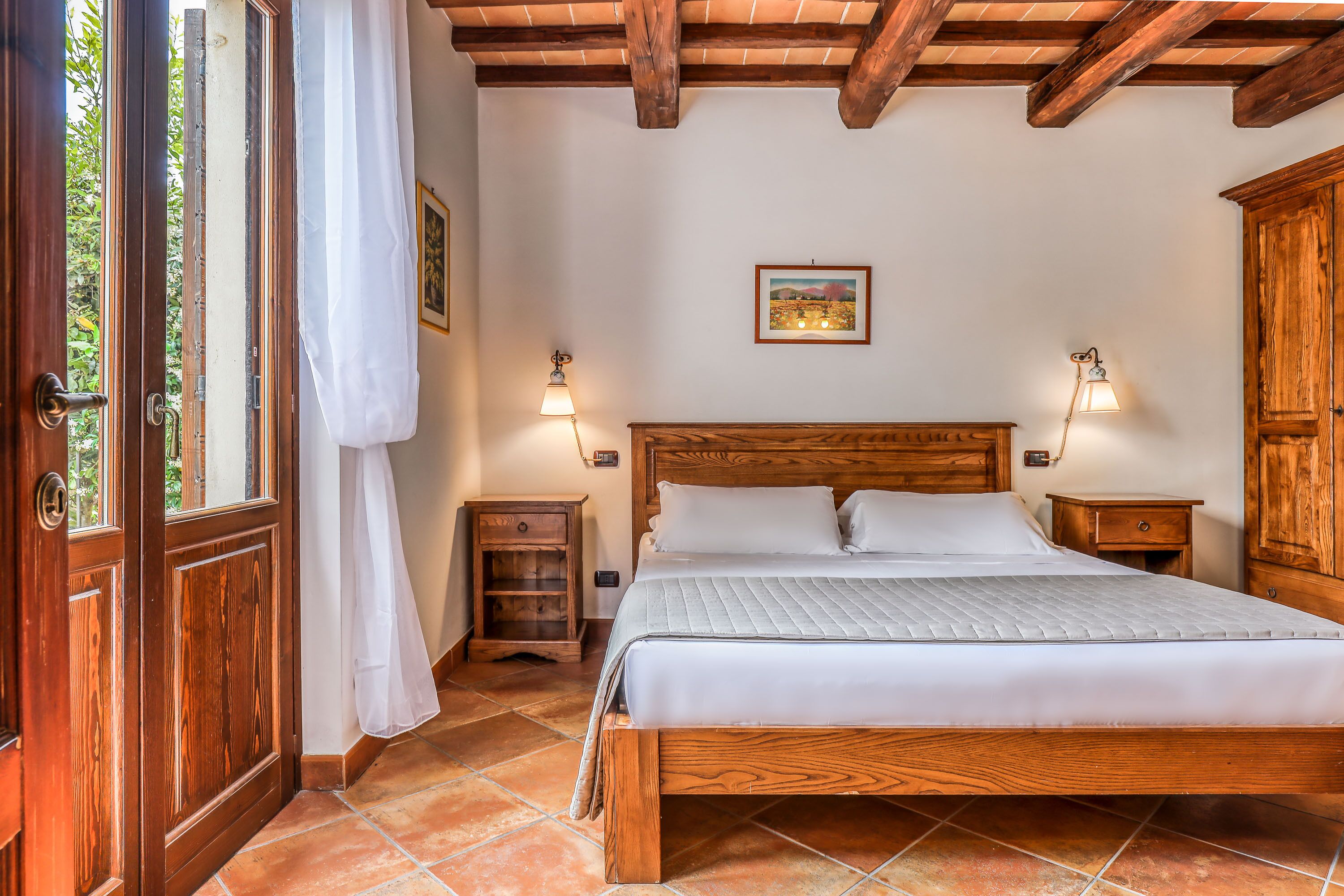 Uma cama de madeira com cabeceira, duas mesas de cabeceira com abajures, um guarda-roupa de madeira e uma janela com cortina.