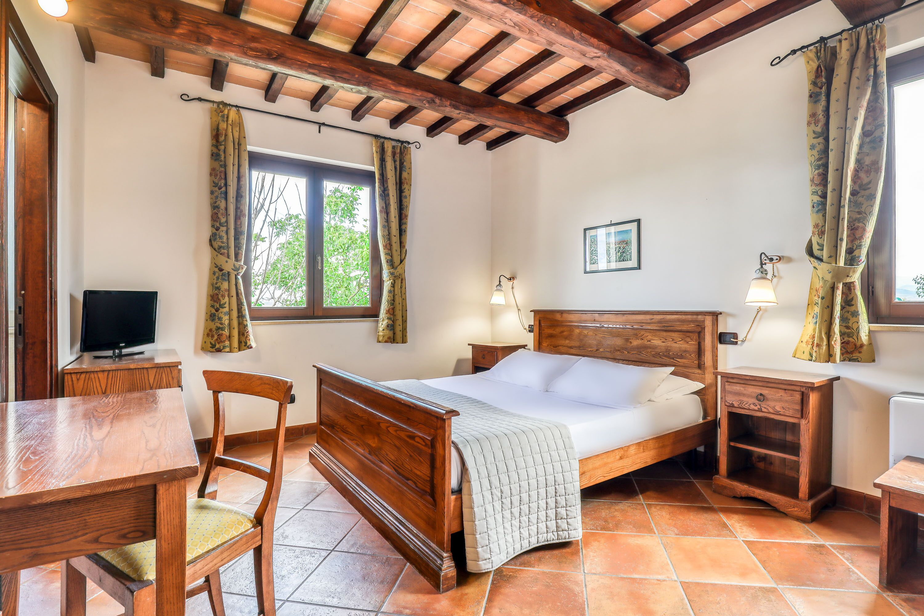 Quarto com cama de madeira, escrivaninha, cadeira e janela com cortinas.