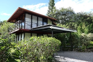 Uma casa de dois andares com varanda, rodeada por muita vegetação.