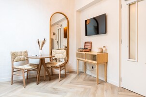Quarto com duas cadeiras de vime, uma mesa redonda de madeira, um espelho grande, um aparador de madeira com uma televisão em cima e uma porta com janela de vidro deslizante.