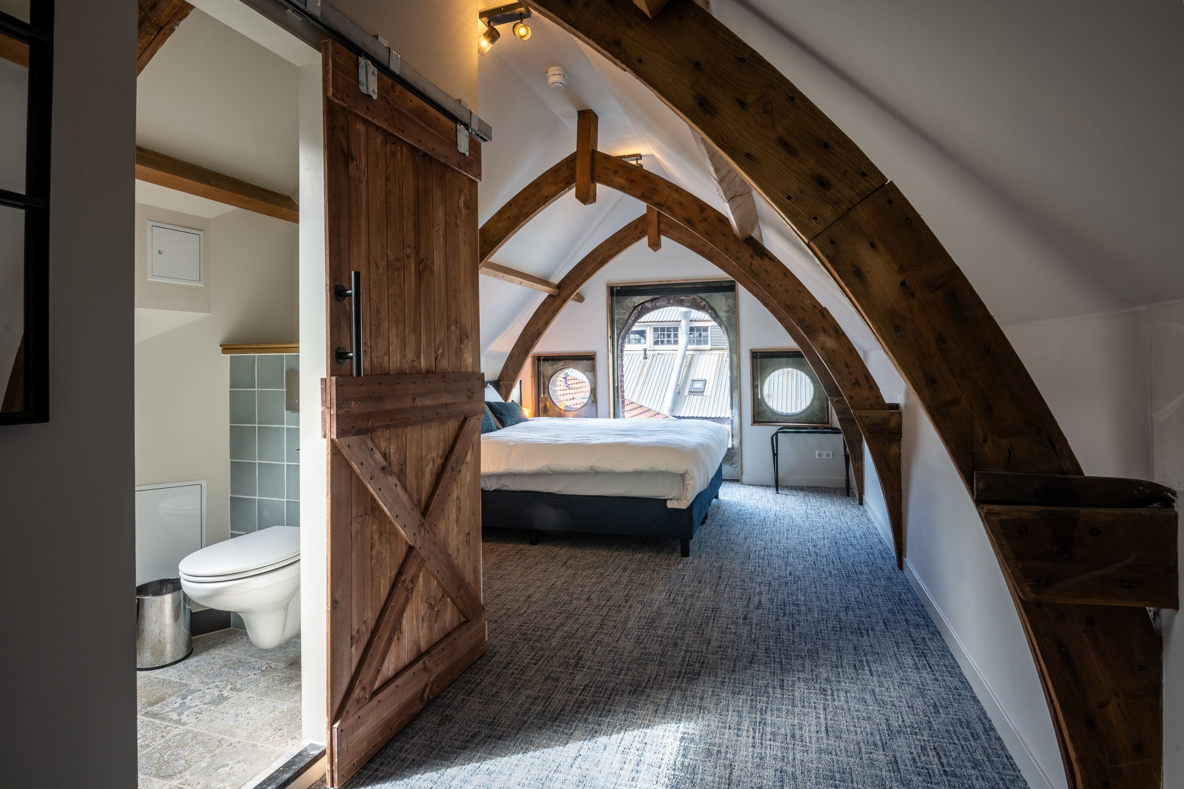 Quarto com teto inclinado, vigas de madeira, cama e vista para o exterior através de uma janela.