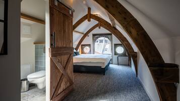 Quarto com teto inclinado, vigas de madeira, cama e vista para o exterior através de uma janela.