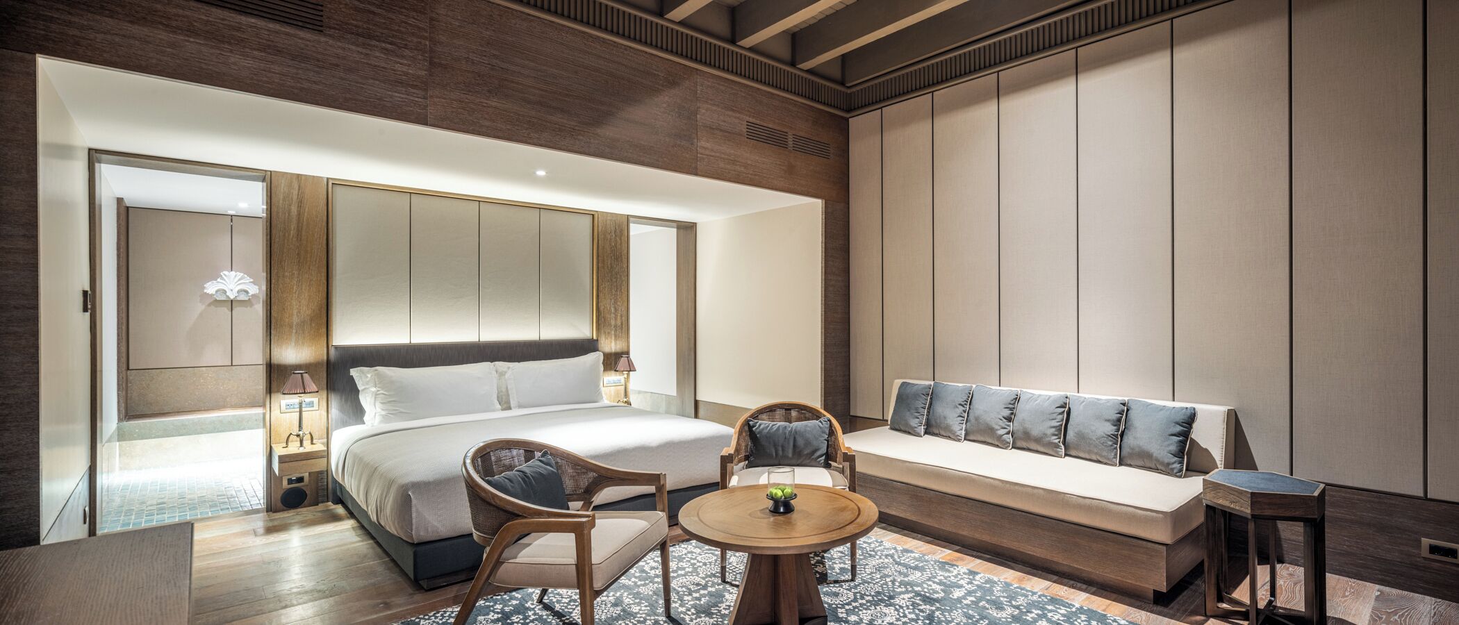 Quarto de hotel moderno com uma cama grande, um sofá, duas cadeiras, uma mesinha e uma janela ampla.