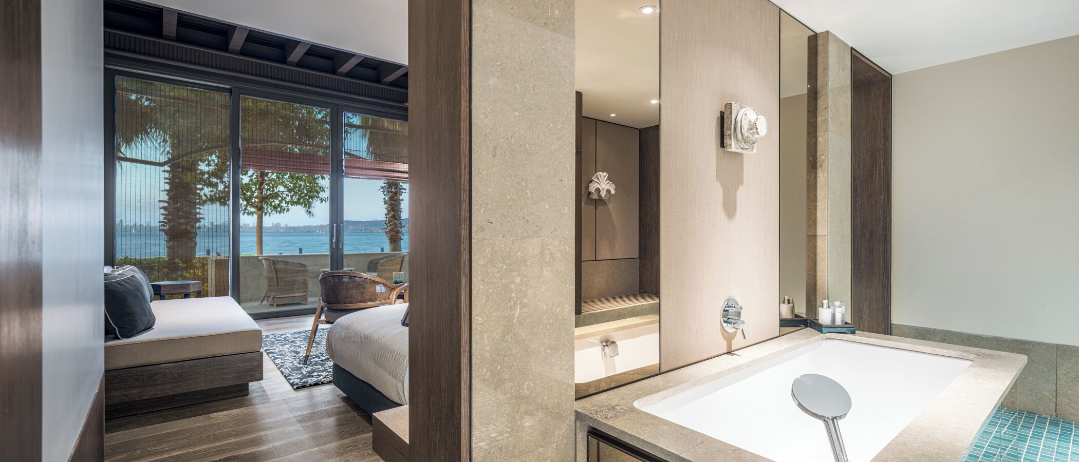 Quarto moderno com uma janela grande, área de estar com sofá e cadeiras, e banheiro com banheira e um espelho grande.