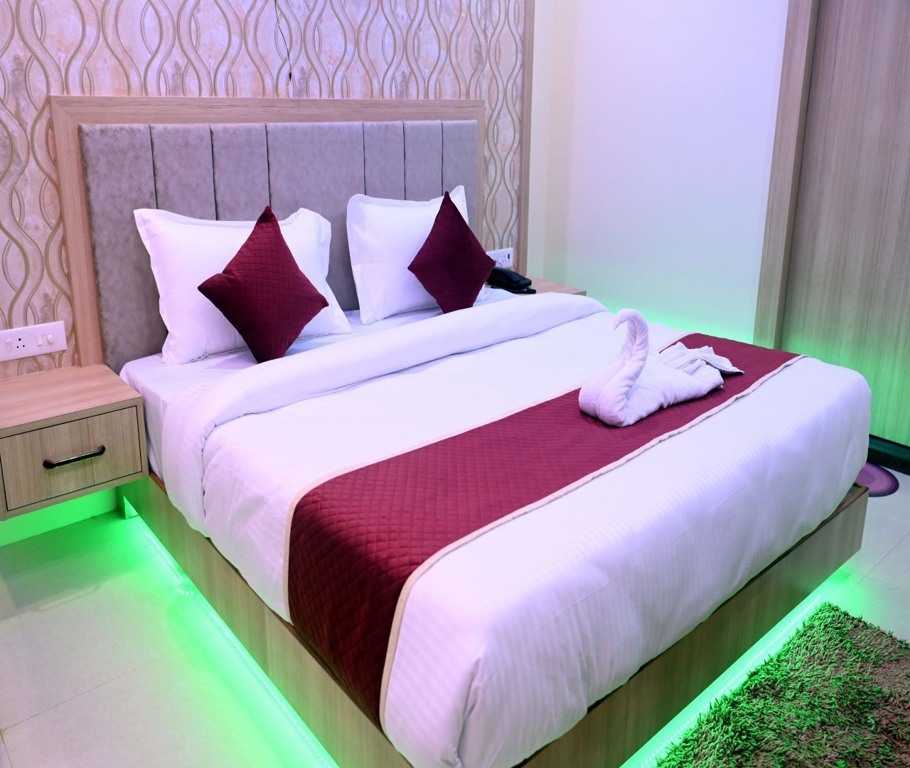 Uma cama bem arrumada com roupa de cama branca e bordô, uma cabeceira de madeira e uma faixa de luz LED verde sob a cama.