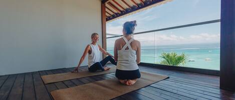 Duas pessoas praticando yoga em tapetes em um quarto com piso de madeira e uma grande janela com vista para o oceano.