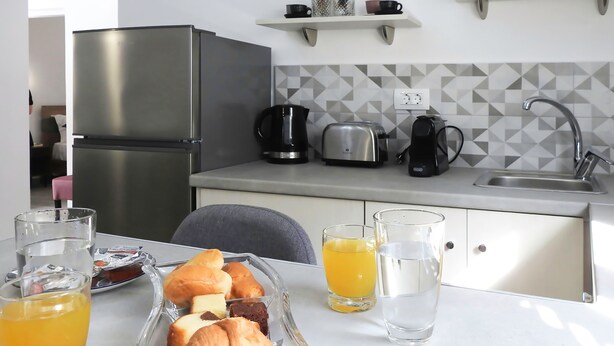 Disposição para o café da manhã com croissants, torradas, suco de laranja e água sobre uma mesa em cozinha moderna com geladeira, torradeira e chaleira.
