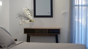 Quarto com cama, mesa de cabeceira com um vaso de flores brancas, espelho na parede e janela com cortinas.