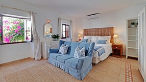Quarto com cama, sofá azul, mesa de cabeceira com abajur, guarda-roupa e janela com cortinas.