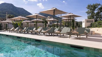 Área de piscina com espreguiçadeiras e guarda-sóis, piscina cristalina e montanhas ao fundo.