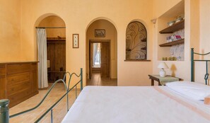 Quarto com cama, cômoda de madeira, portas em arco e uma mesinha com abajur.