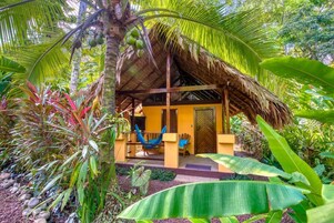 Uma cabana com telhado de palha, uma rede e porta de madeira, cercada por vegetação tropical exuberante.
