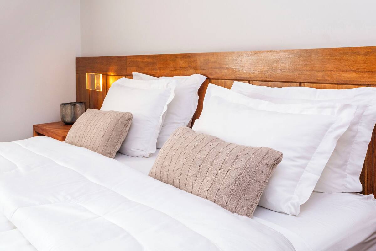 Cama bem arrumada com roupa de cama branca e duas almofadas bege, uma cabeceira de madeira e uma mesa de cabeceira com abajur.