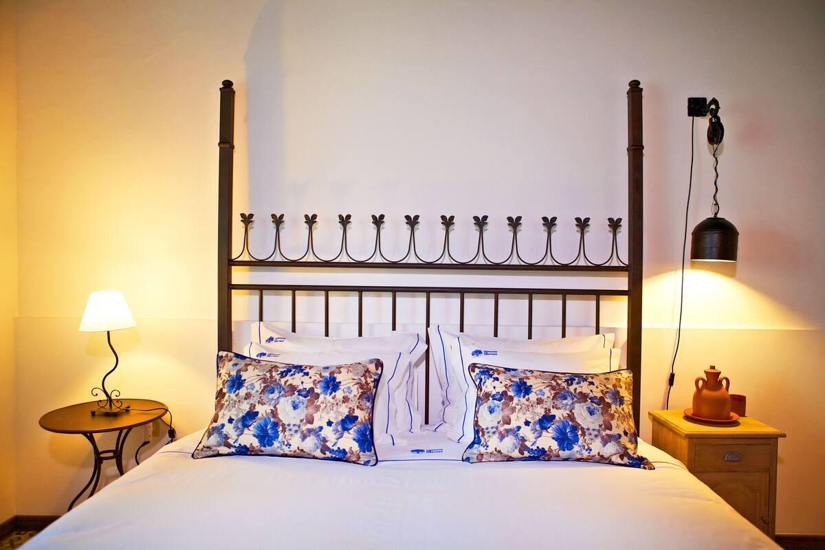 Cama bem arrumada com almofadas florais, mesa de cabeceira com abajur e criado-mudo com abajur e um vaso.