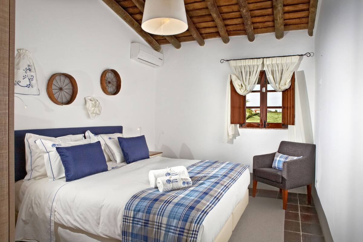 Quarto com cama, cadeira, janela com cortinas e detalhes decorativos nas paredes.