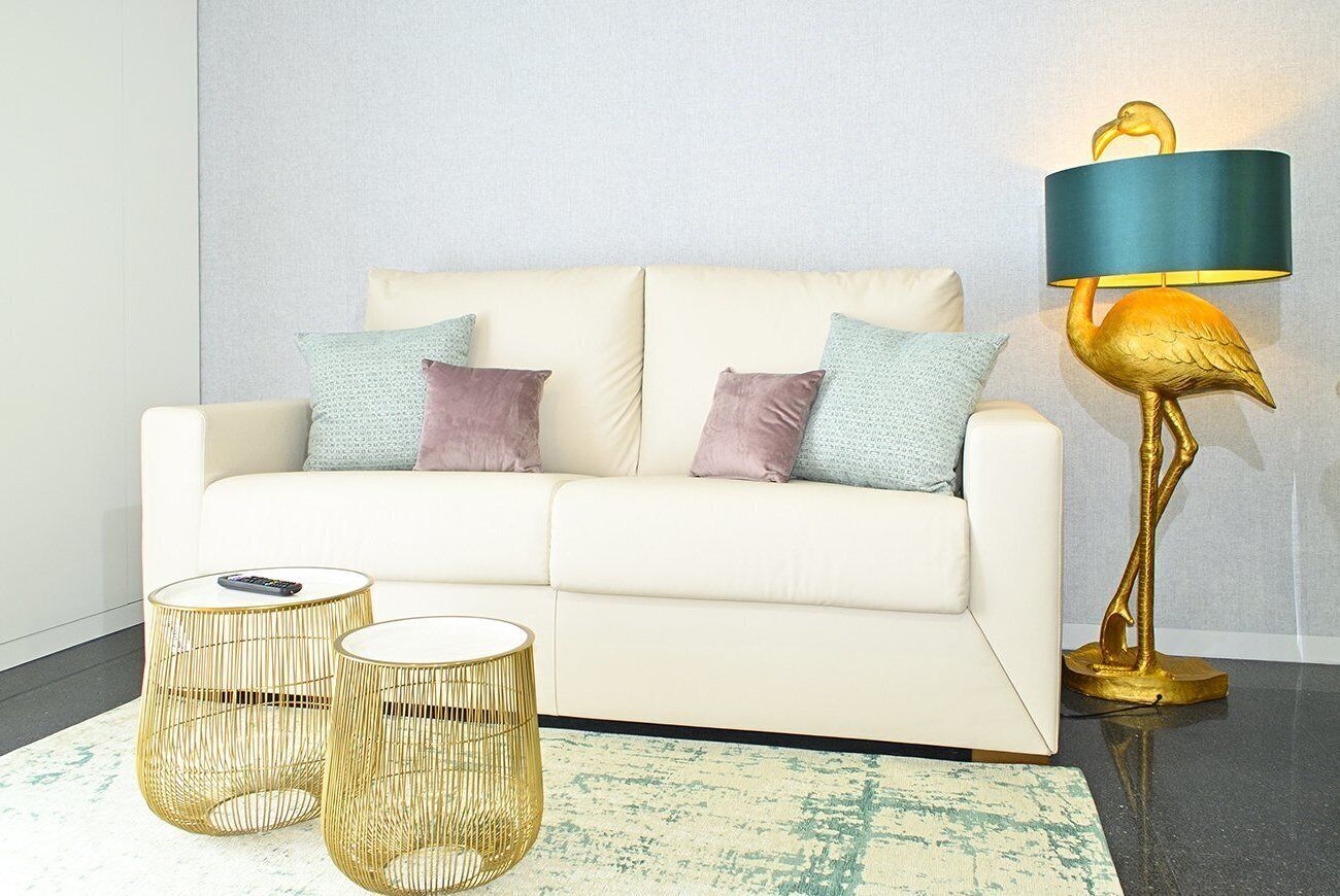 Um sofá branco com duas almofadas, duas mesinhas de apoio com detalhes dourados e um candeeiro de chão dourado com um abajur azul.