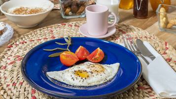 Mesa de café da manhã com um prato azul de ovos, tomates e azeitonas, uma cadeira de vime e vista para o mar.