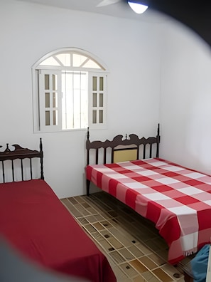 Quarto pequeno com uma cama de solteiro, coberta por um edredom xadrez vermelho e branco, uma janela em arco branca e parede branca.