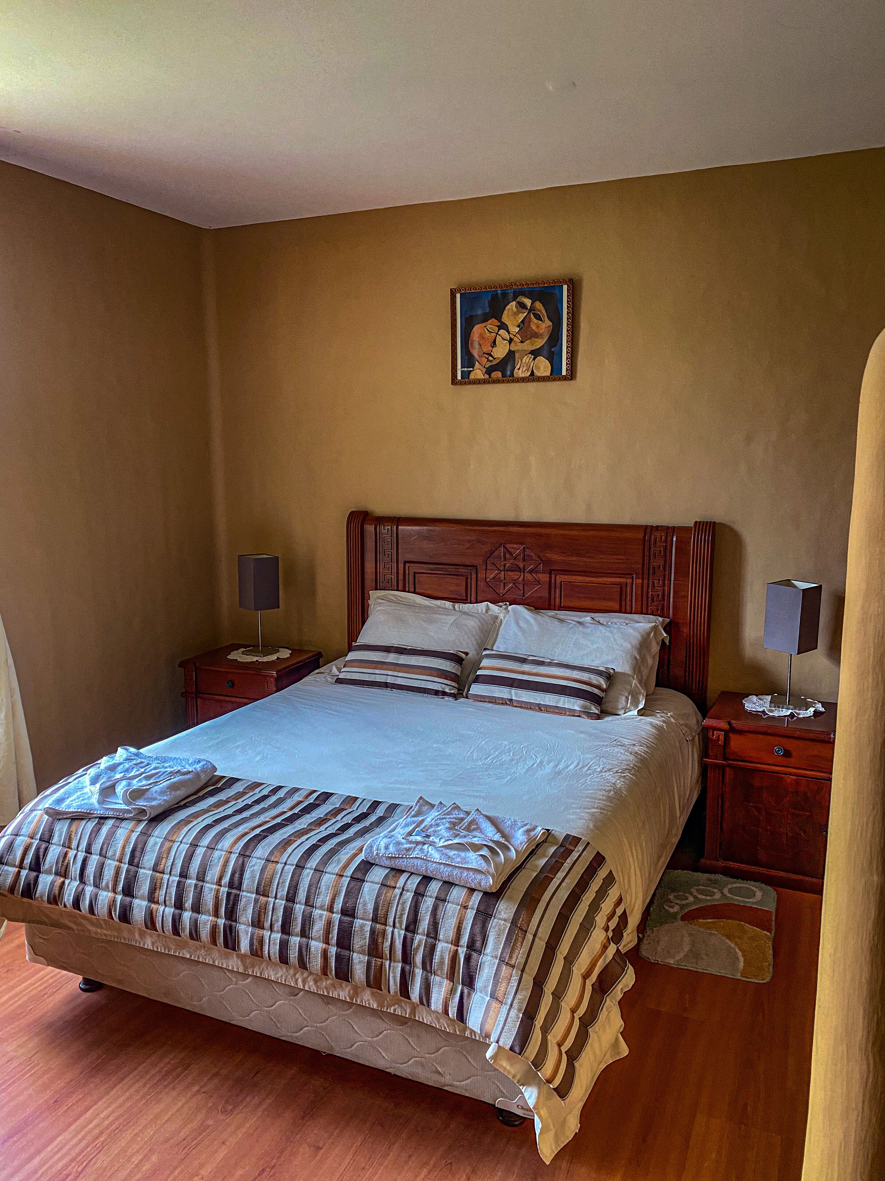 Uma cama bem arrumada com roupa de cama listrada, duas mesas de cabeceira com abajures, uma cabeceira de madeira e um quadro na parede.